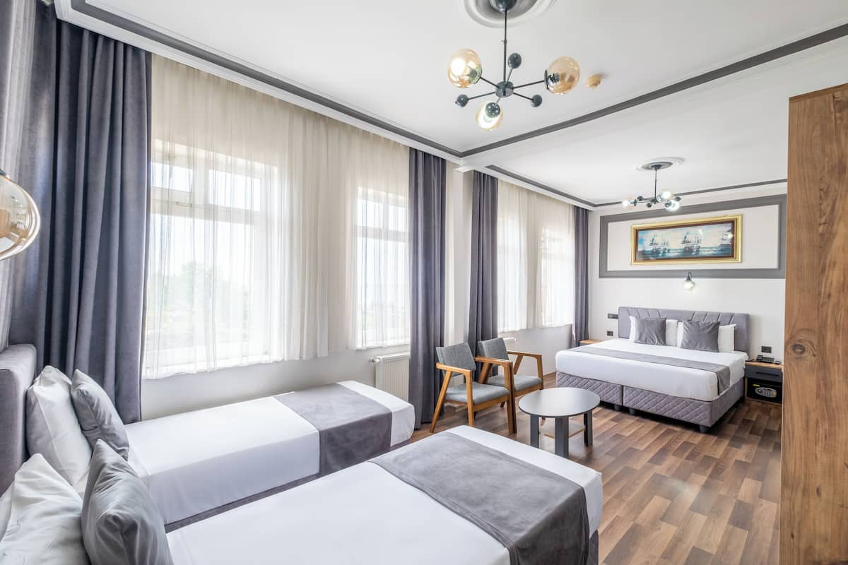 Quarto de hotel com duas camas, uma escrivaninha, uma cadeira e uma janela grande com cortinas.