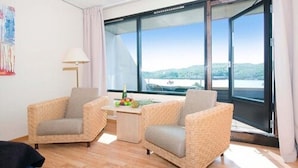 Quarto com duas poltronas de vime, uma mesa com frutas e vista para um lago através de grandes janelas.