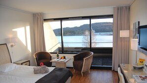 Quarto de hotel com cama, uma cadeira, uma mesinha com garrafa e frutas, e uma janela grande com vista para um corpo d'água e colinas.