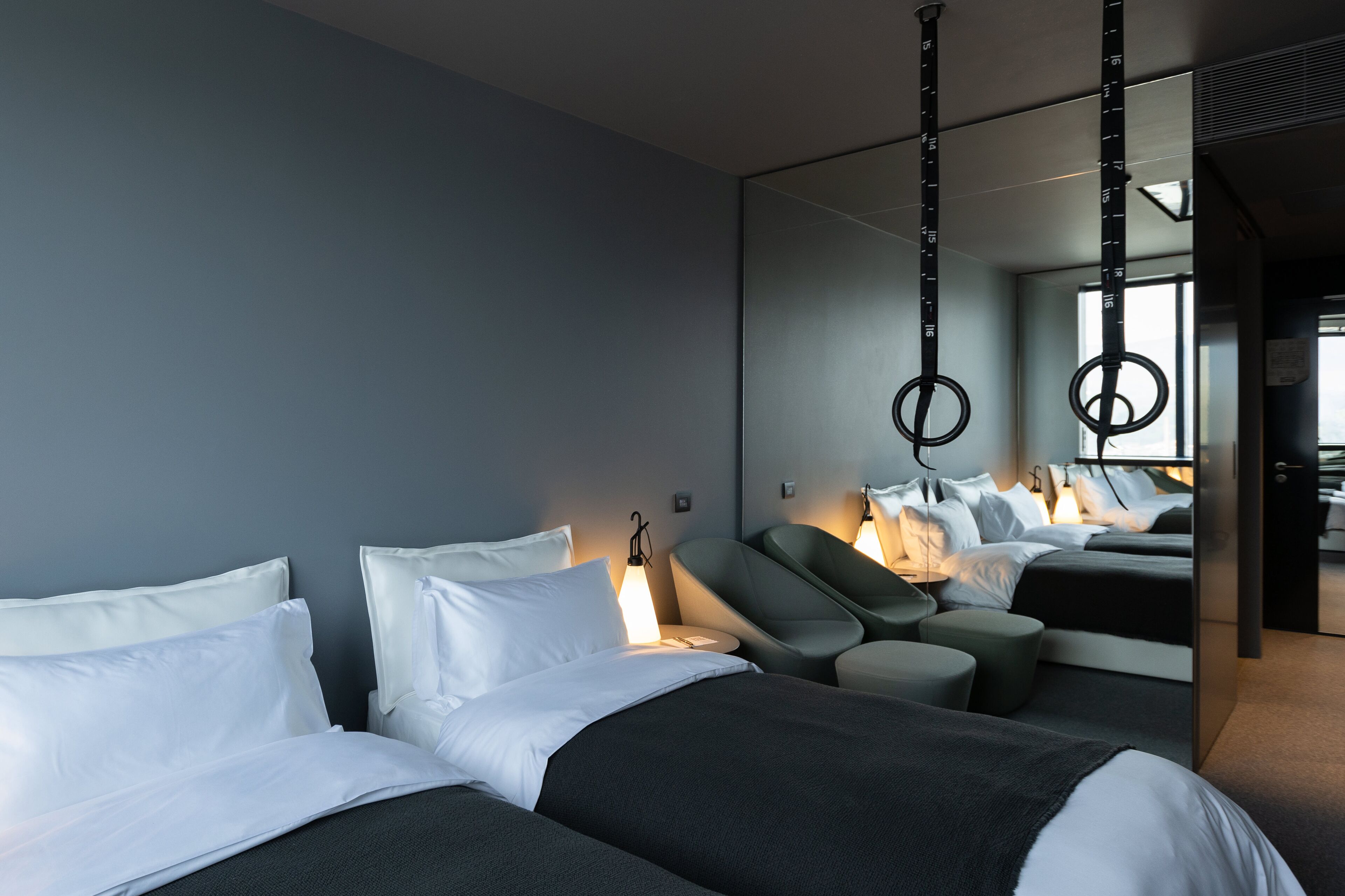 Quarto de hotel moderno com duas camas, área de estar e um espelho grande que reflete o quarto.