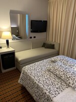 Quarto de hotel com uma cama, uma mesinha de cabeceira com abajur, televisão na parede e um mini frigorífico.