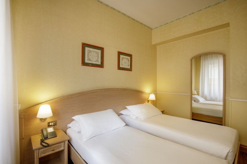 Quarto de hotel com duas camas de solteiro, mesa de cabeceira com abajur, telefone e espelho.