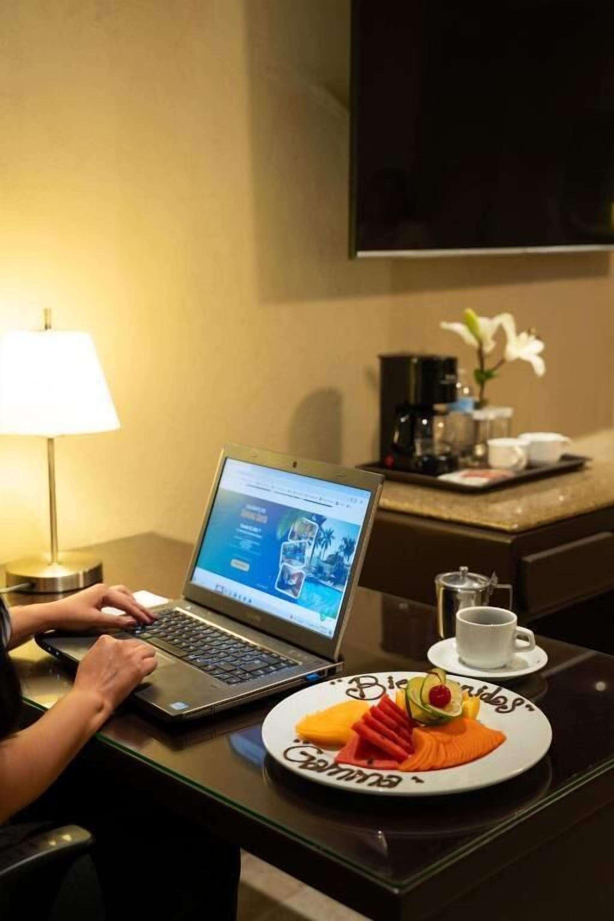 Um laptop sobre uma mesa, com uma travessa de fruta e uma chávena de café, uma lâmpada e uma máquina de café ao fundo.