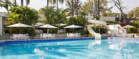 Piscina com espreguiçadeiras e guarda-sóis, cercada por palmeiras e um tobogã espiral branco.