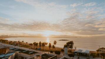 Vista costeira com edifícios, palmeiras e um pôr do sol sobre o oceano.