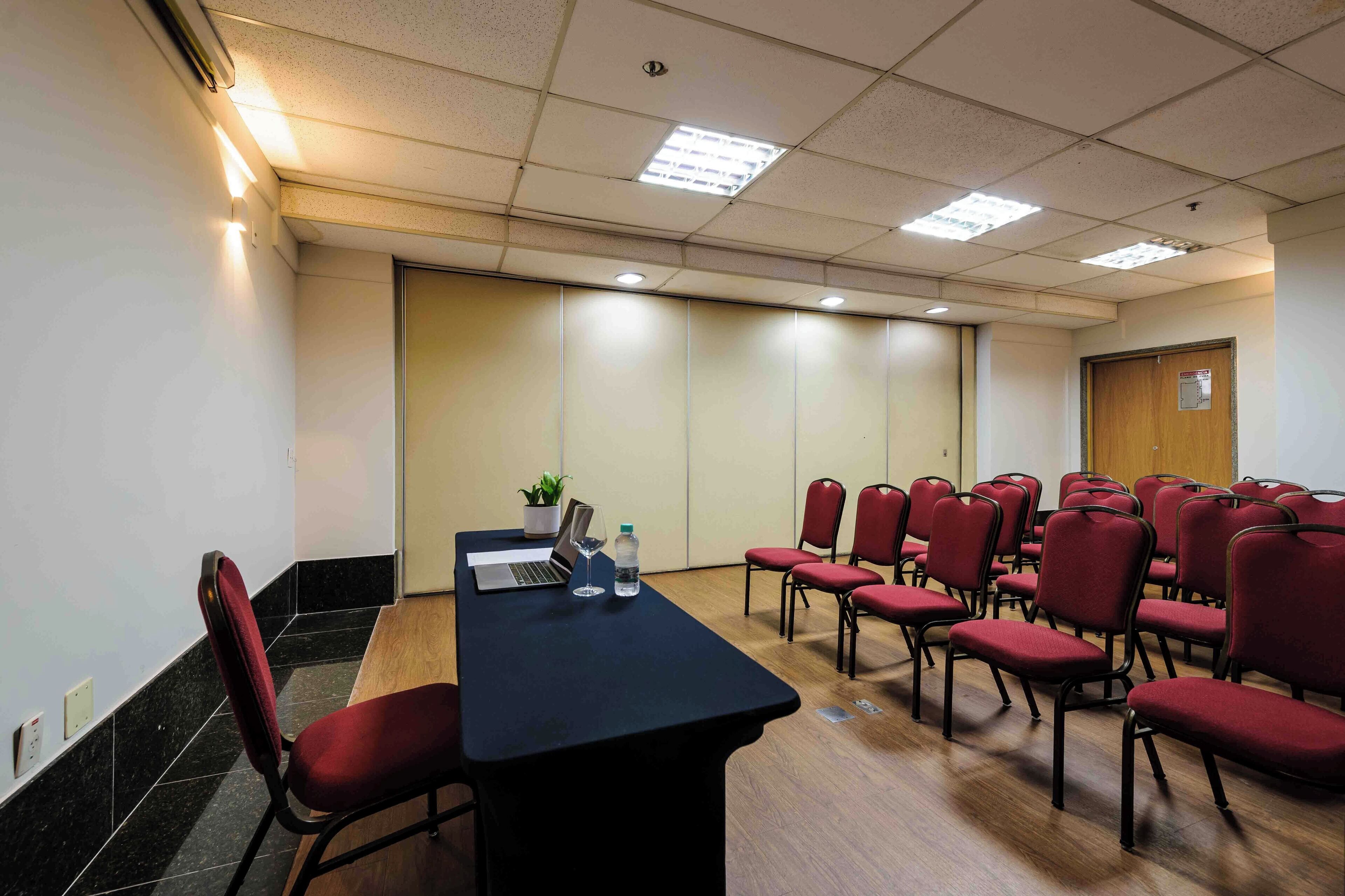 Uma sala de conferências com uma mesa comprida, cadeiras vermelhas, um laptop, uma planta e uma garrafa de água.