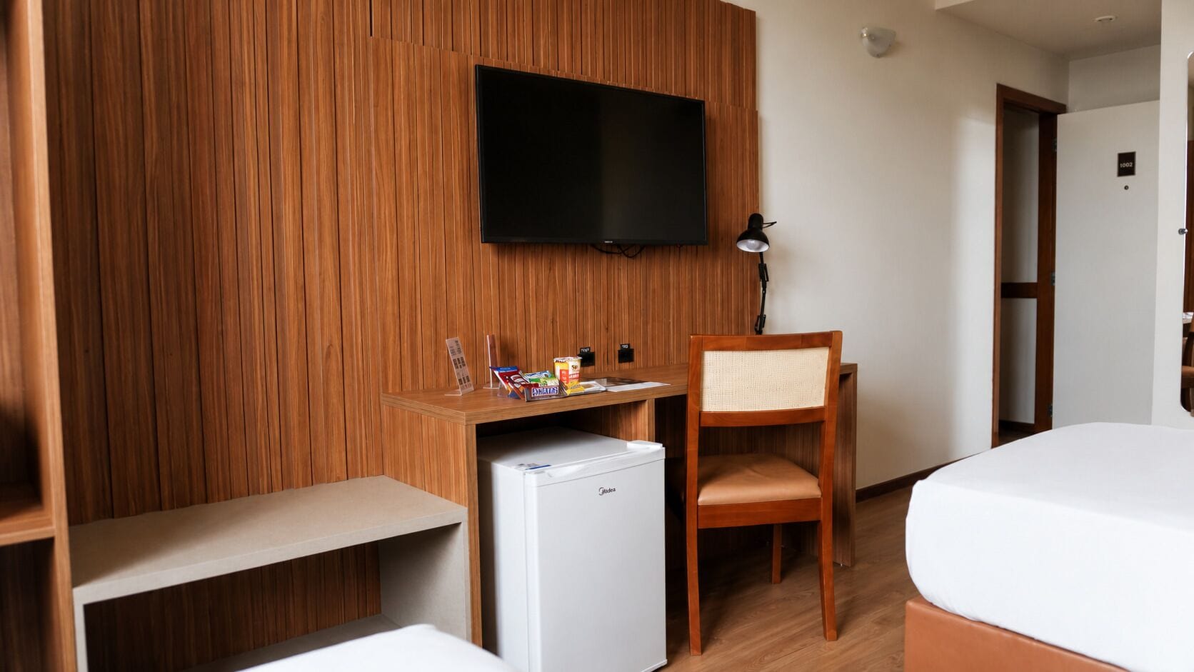 Quarto de hotel com parede de painel de madeira, televisão de tela plana fixada acima de uma escrivaninha, mini-geladeira e uma cadeira de madeira com assento acolchoado, além de uma cama com roupa de cama branca.