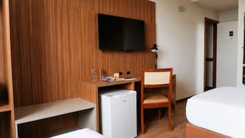 Quarto de hotel com parede de painel de madeira, televisão de tela plana fixada acima de uma escrivaninha, mini-geladeira e uma cadeira de madeira com assento acolchoado, além de uma cama com roupa de cama branca.