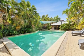 Uma piscina retangular com água cristalina, rodeada por vegetação tropical exuberante e uma estrutura coberta ao fundo.
