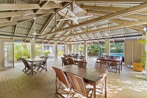 Uma área de refeições coberta ao ar livre, com mesas e cadeiras, um ventilador de teto e vista para a piscina.