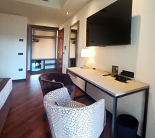 Quarto de hotel com cama, escrivaninha, televisão, cadeira e lixeira.