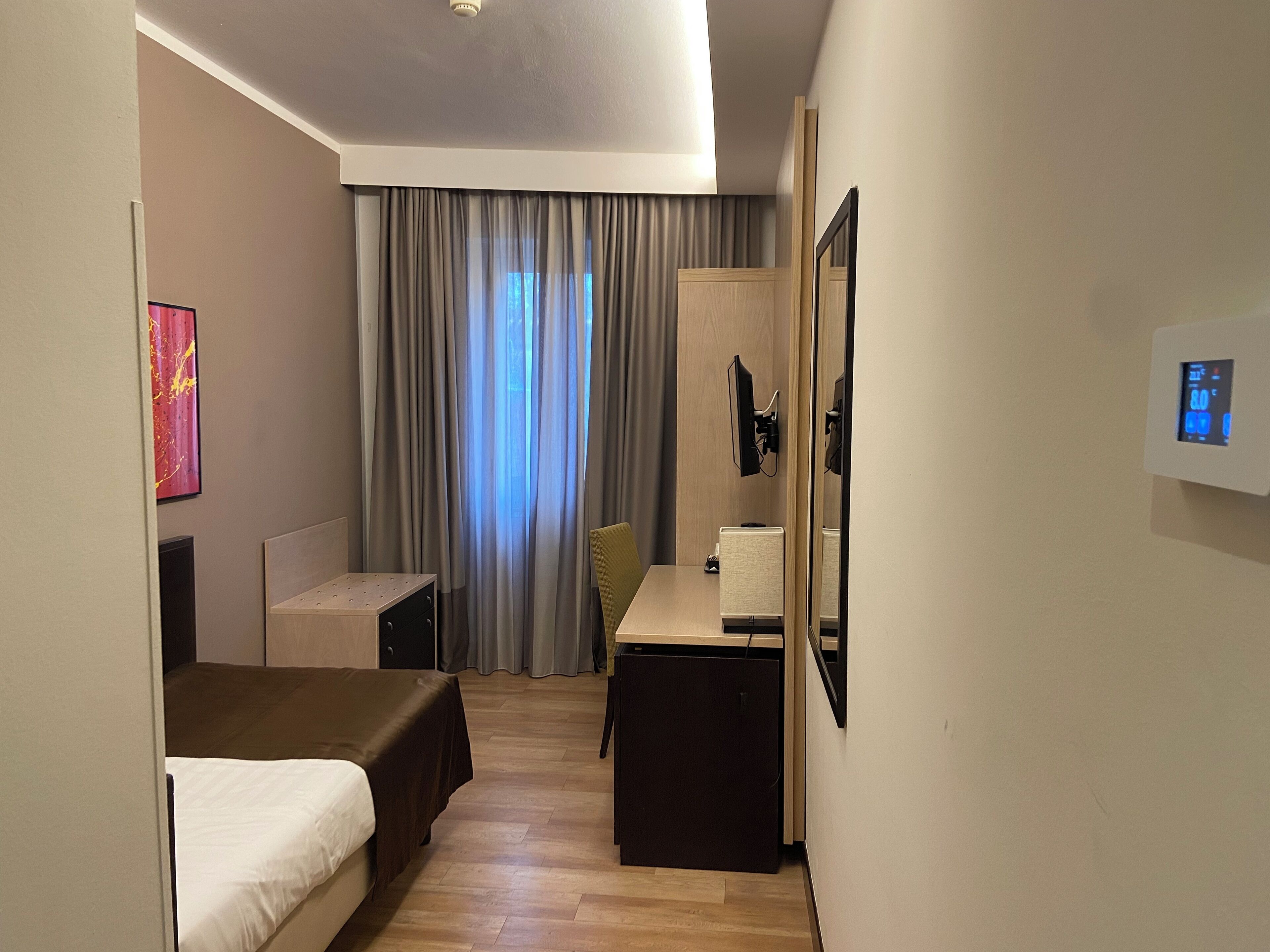 Quarto de hotel com cama, escrivaninha, cadeira, televisão e termostato.