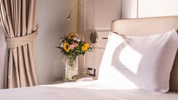 Cama bem arrumada com lençóis brancos, um vaso com flores, abajur e um telefone em uma mesinha de cabeceira.