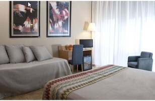 Quarto de hotel com cama, escrivaninha com cadeira, abajur e duas pinturas abstratas na parede.