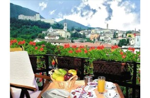 Uma mesa com uma cesta de frutas, um copo de sumo de laranja e uma chávena numa varanda com vista para uma cidade com edifícios históricos e uma montanha ao fundo.