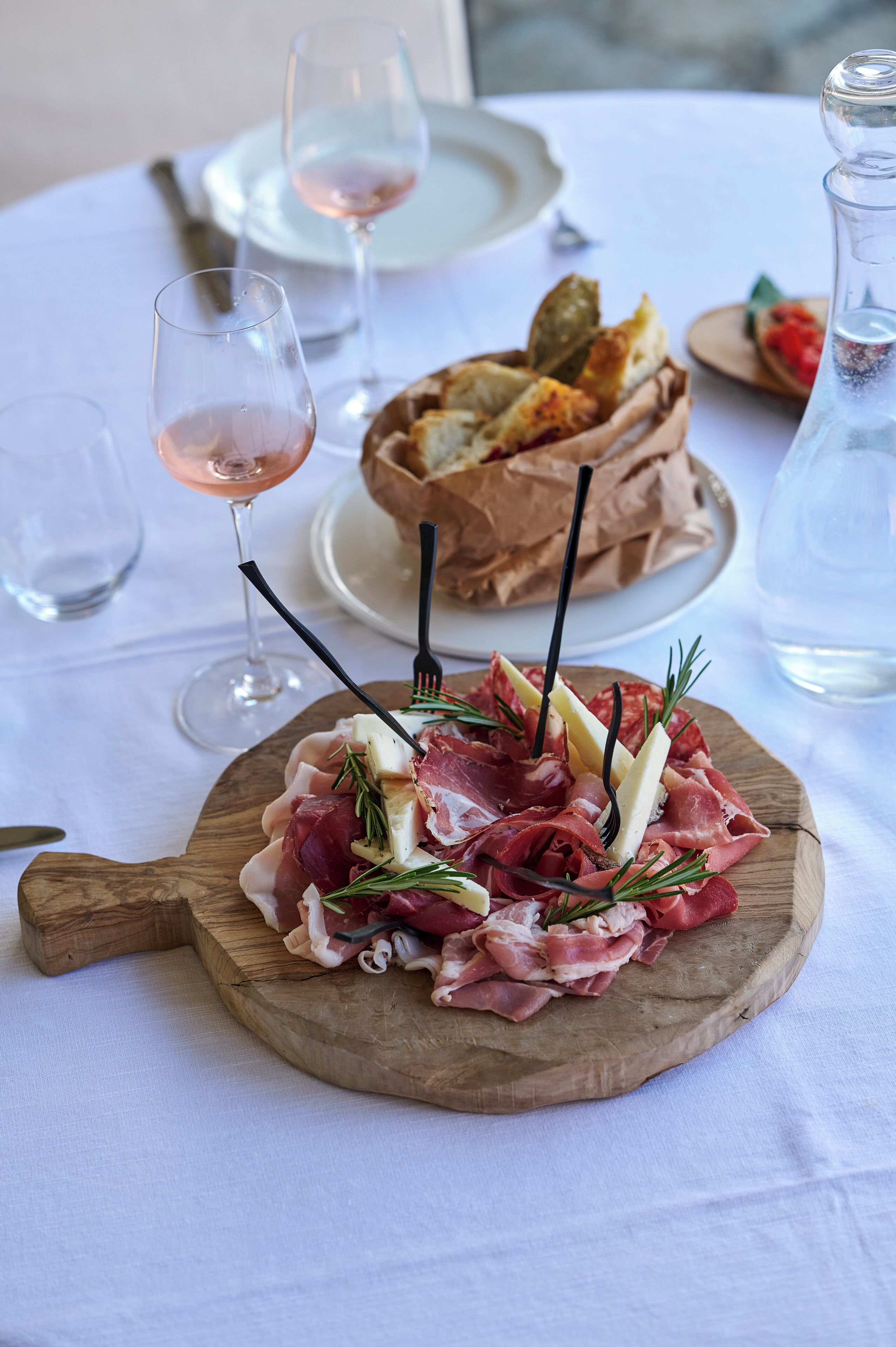 Tábua de madeira com fatias de carne e queijo, acompanhada por um copo de vinho rosé, sobre uma mesa com toalha branca.