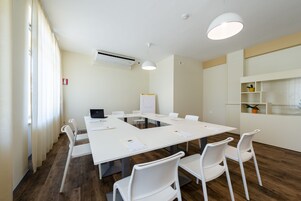 Uma sala de conferências com uma mesa branca, cadeiras brancas, um quadro branco e uma luminária branca no teto.