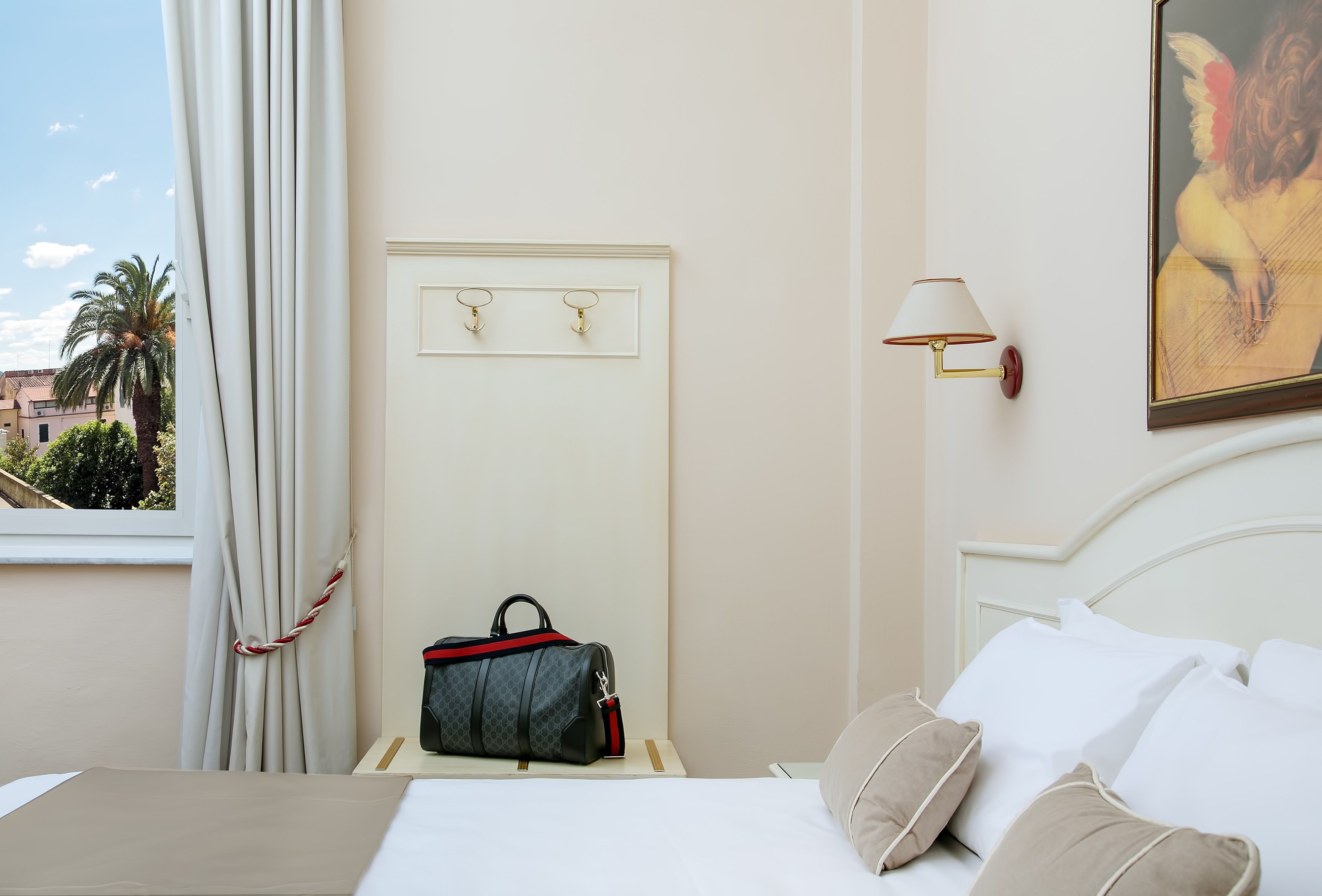 Quarto de hotel com uma cama, mesa de cabeceira, abajur, quadro e vista para palmeiras lá fora.