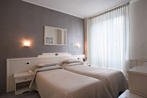 Quarto de hotel com duas camas, uma mesa de cabeceira com telefone, um criado-mudo, um espelho e uma janela com cortinas.