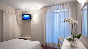 Quarto de hotel com cama, televisão na parede, secretária com planta e janela com cortinas leves.