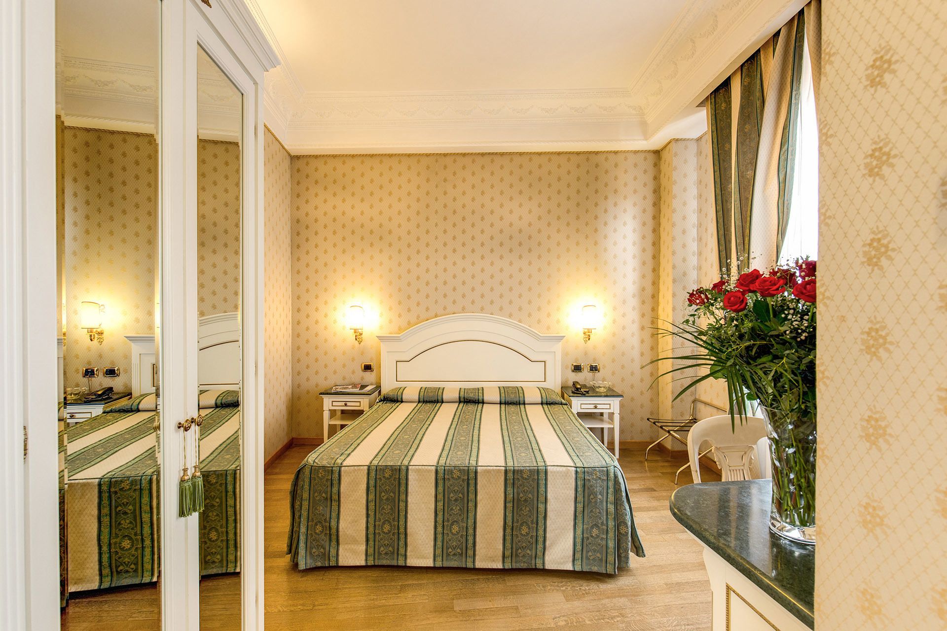 Quarto de hotel com uma cama grande, duas mesas de cabeceira, um espelho e um vaso com flores.
