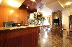 Uma área de recepção de hotel com balcão de madeira, mesa de atendimento, um pilar de mármore e um espaço de refeições com cadeiras e uma mesa.