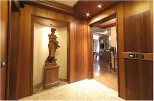 Corredor interno de madeira com uma estátua em pedestal, uma porta e um vislumbre de uma sala de jantar com mesa e cadeiras.