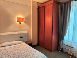 Quarto de hotel com cama, mesa de cabeceira, guarda-roupa de madeira, luminária na parede e janela com cortinas.