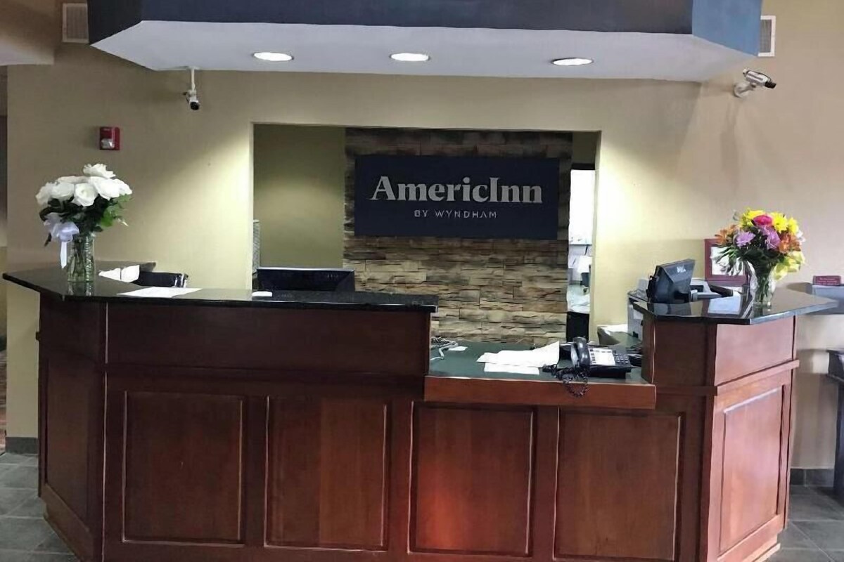 Bancada de recepção com uma placa indicando 'AmericInn by Wyndham', uma parede de pedra e um vaso com flores.