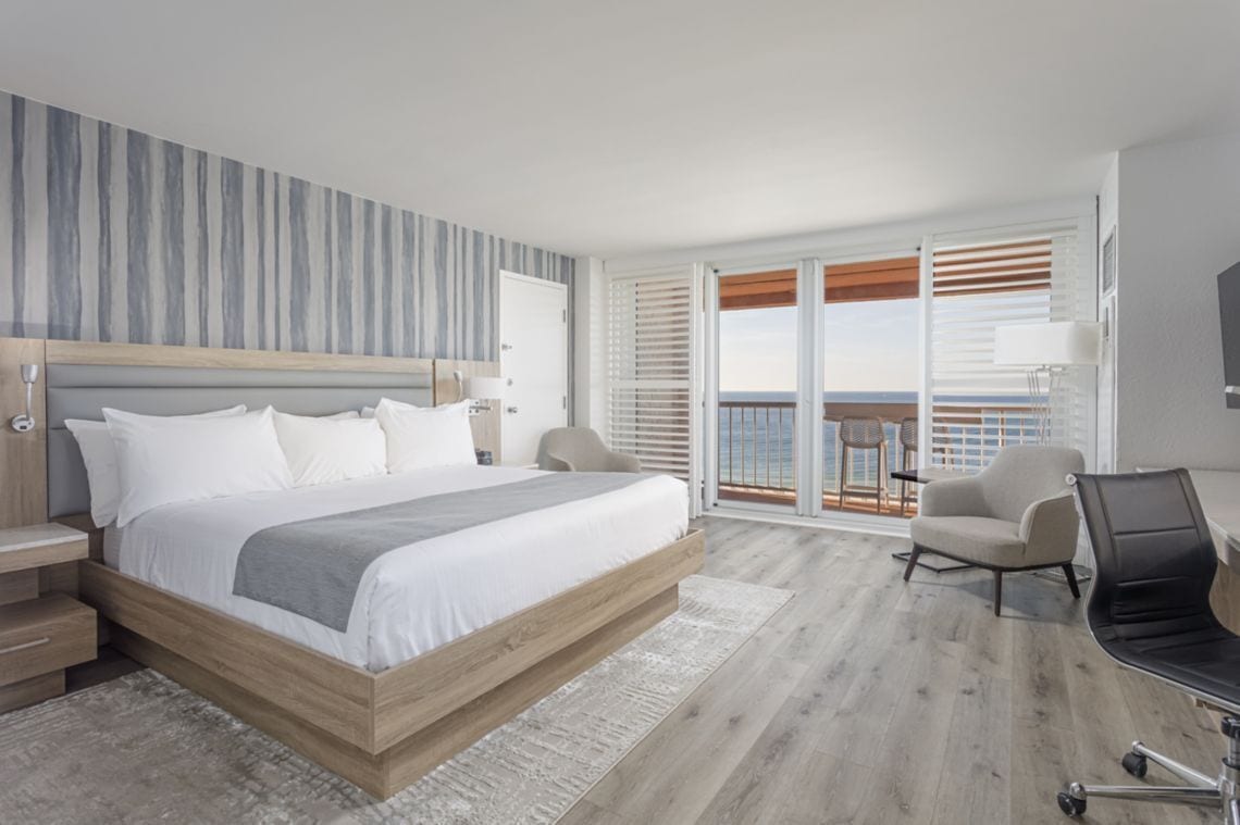 Quarto de hotel com cama, uma cadeira, escrivaninha, varanda com vista e uma janela grande.