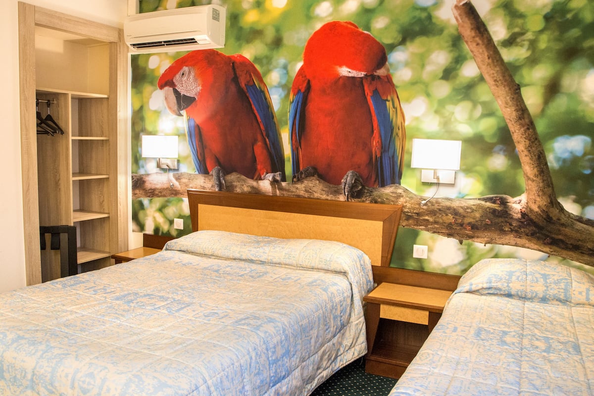 Quarto de hotel com duas camas, cabeceira de madeira e um mural de araras na parede.