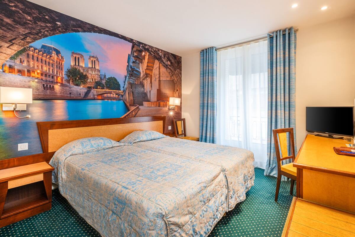 Quarto de hotel com uma cama grande, uma escrivaninha, uma cadeira, uma TV e um mural de um edifício histórico.
