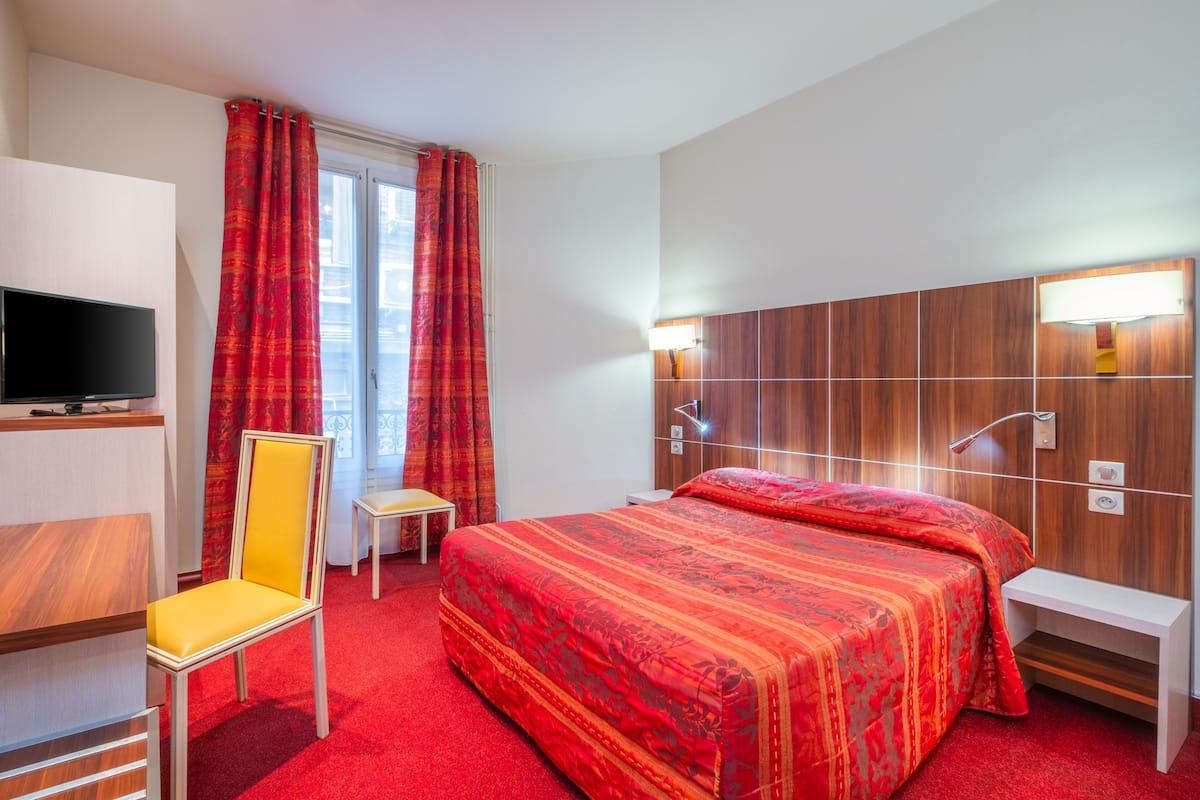 Quarto de hotel com cama, televisão, cadeira, escrivaninha e janela com cortinas.