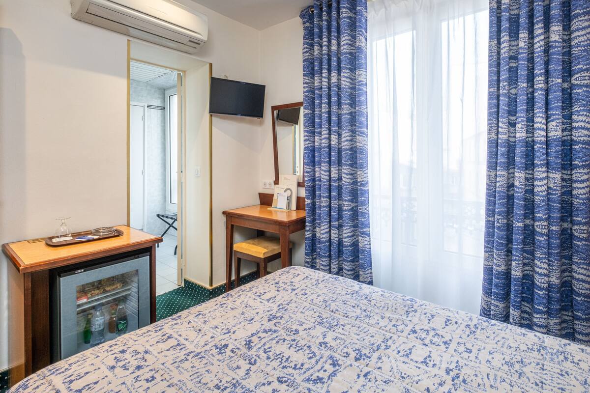 Quarto de hotel com cama, escrivaninha, televisão, minibar e janela com cortinas.