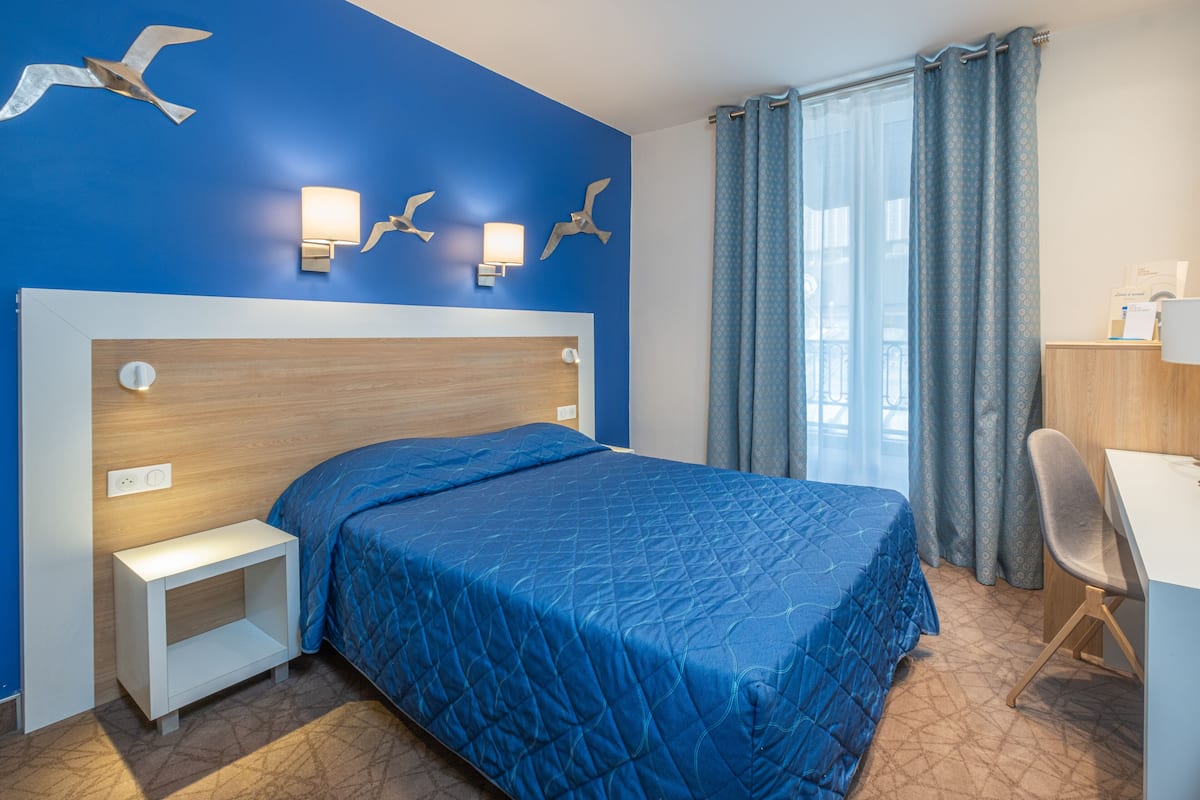 Quarto de hotel com uma cama grande, uma escrivaninha e uma cadeira. Há arandelas e uma janela com cortinas.