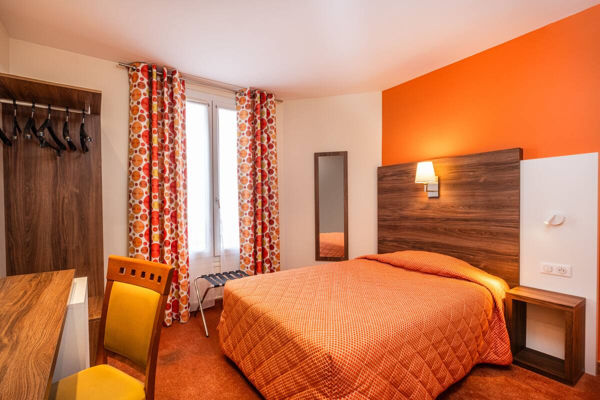 Quarto de hotel com cama, cabeceira de madeira, mesa de cabeceira, cadeira, escrivaninha e janela com cortinas.