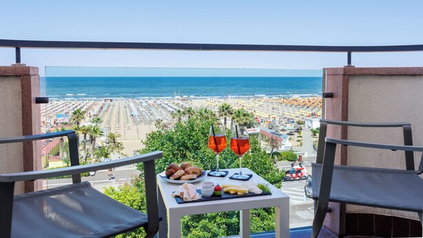 Varanda com mesa posta para refeição, com vista para uma praia com guarda-sóis e céu claro.