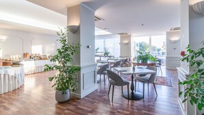 Uma ampla área de refeições com piso de madeira, paredes brancas e várias mesas e cadeiras. Há plantas em vasos e uma mesa de buffet com diversos alimentos.