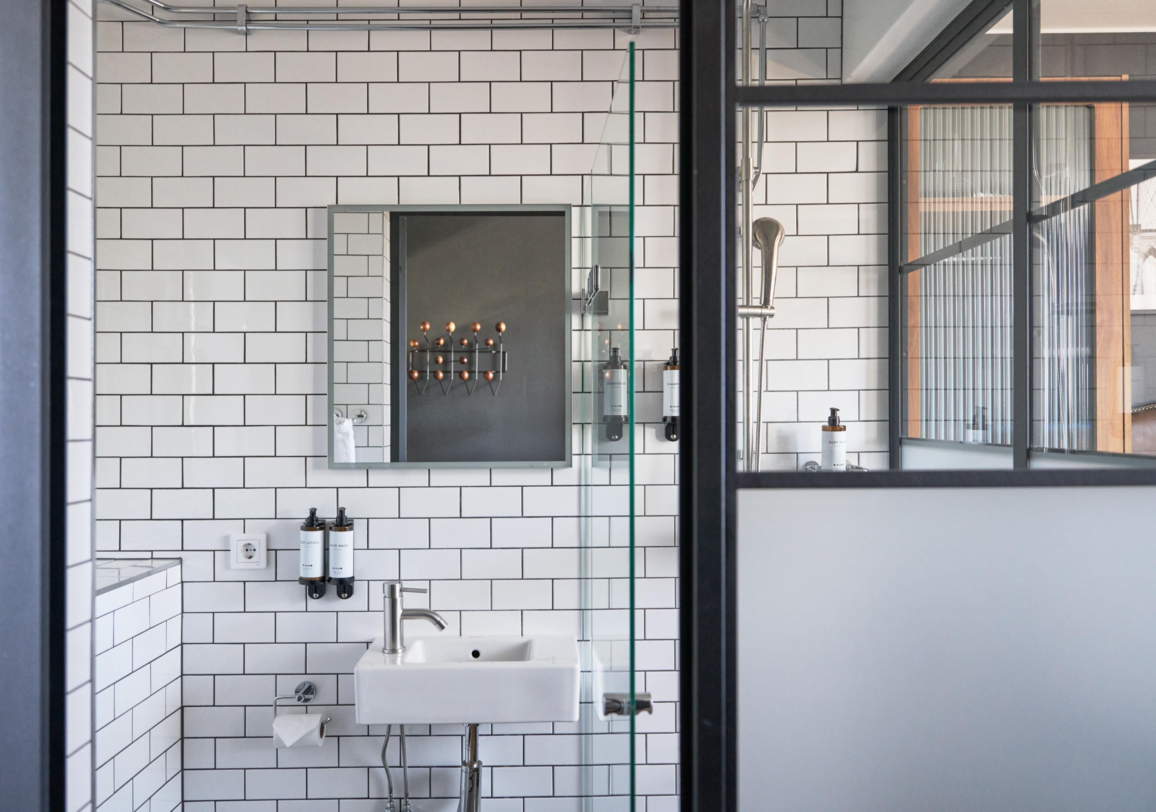 Um banheiro moderno com parede de azulejos brancos, box de vidro, pia branca e espelho com porta-toalhas.