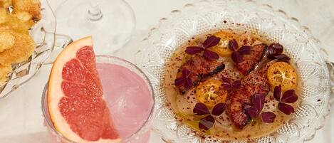 Uma mesa com diversos alimentos e bebidas, incluindo uma taça de vinho branco, uma bebida rosa com uma fatia de toranja, um prato com carne e cítricos, e uma tigela de azeitonas.
