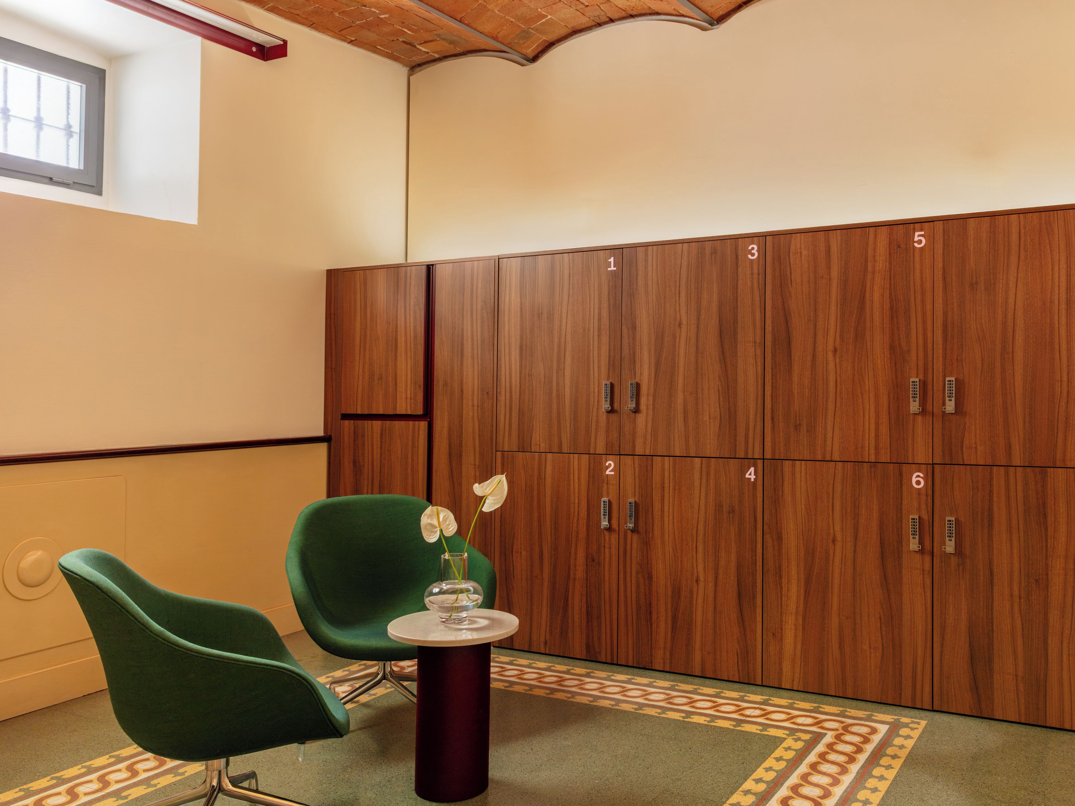 Quarto com uma cadeira verde, uma mesa pequena com um vaso e um guarda-roupa de madeira com divisórias numeradas.