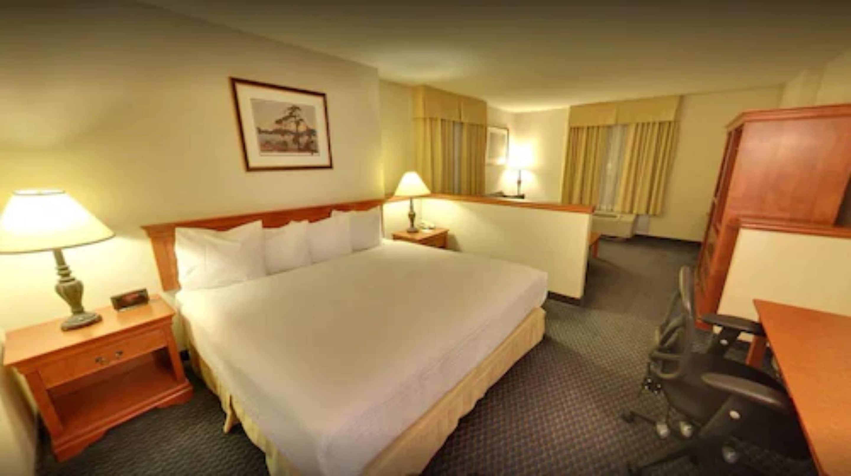 Quarto de hotel com uma cama grande, duas mesas de cabeceira com luminárias, uma escrivaninha com cadeira, um espelho e um quadro na parede.