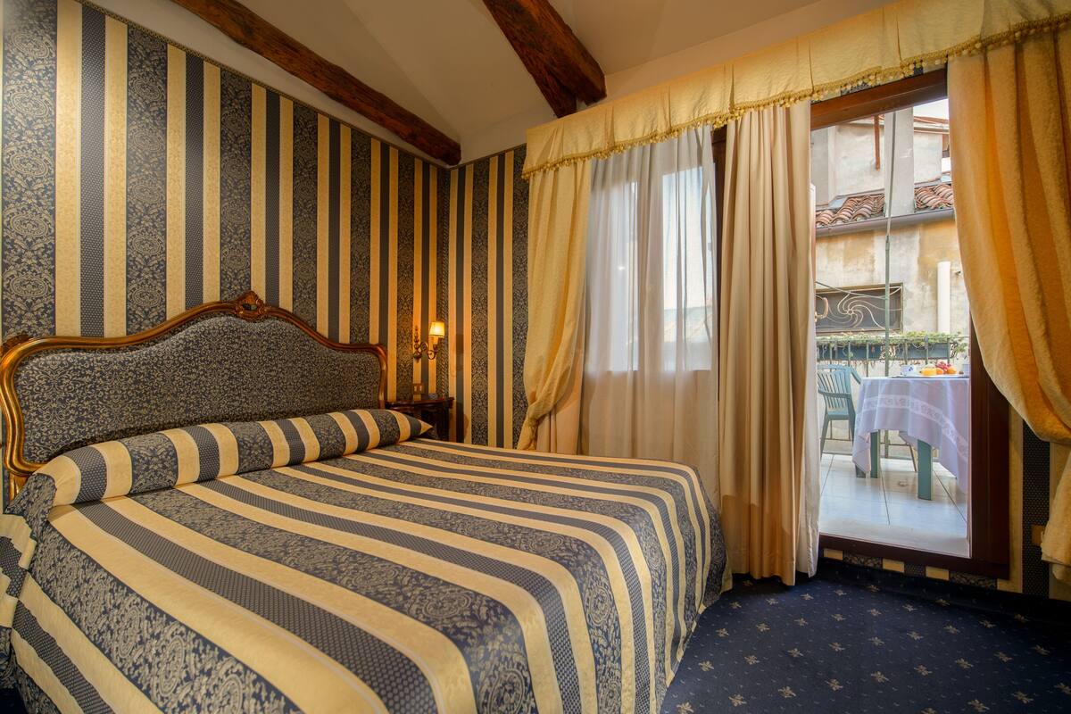 Quarto com cama, janela com cortinas e vista para uma mesa e cadeiras ao ar livre.