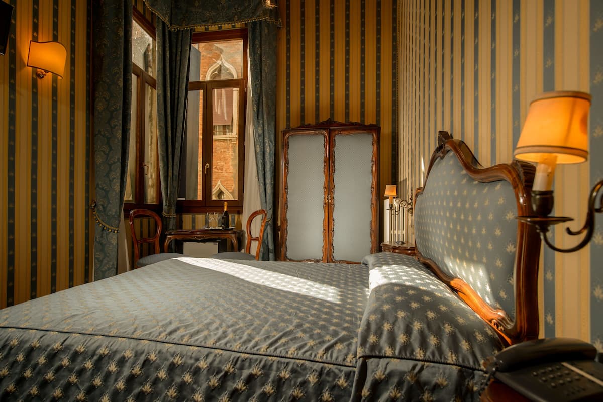 Quarto de hotel com uma cama grande, um guarda-roupa de madeira, uma escrivaninha com cadeira e um telefone.