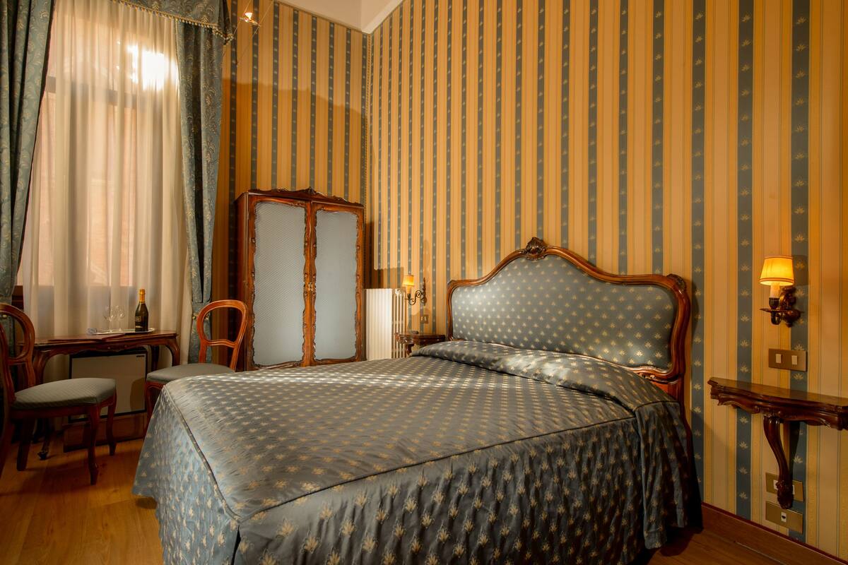 Quarto de hotel com uma cama, uma escrivaninha, uma cadeira, um espelho e um abajur.