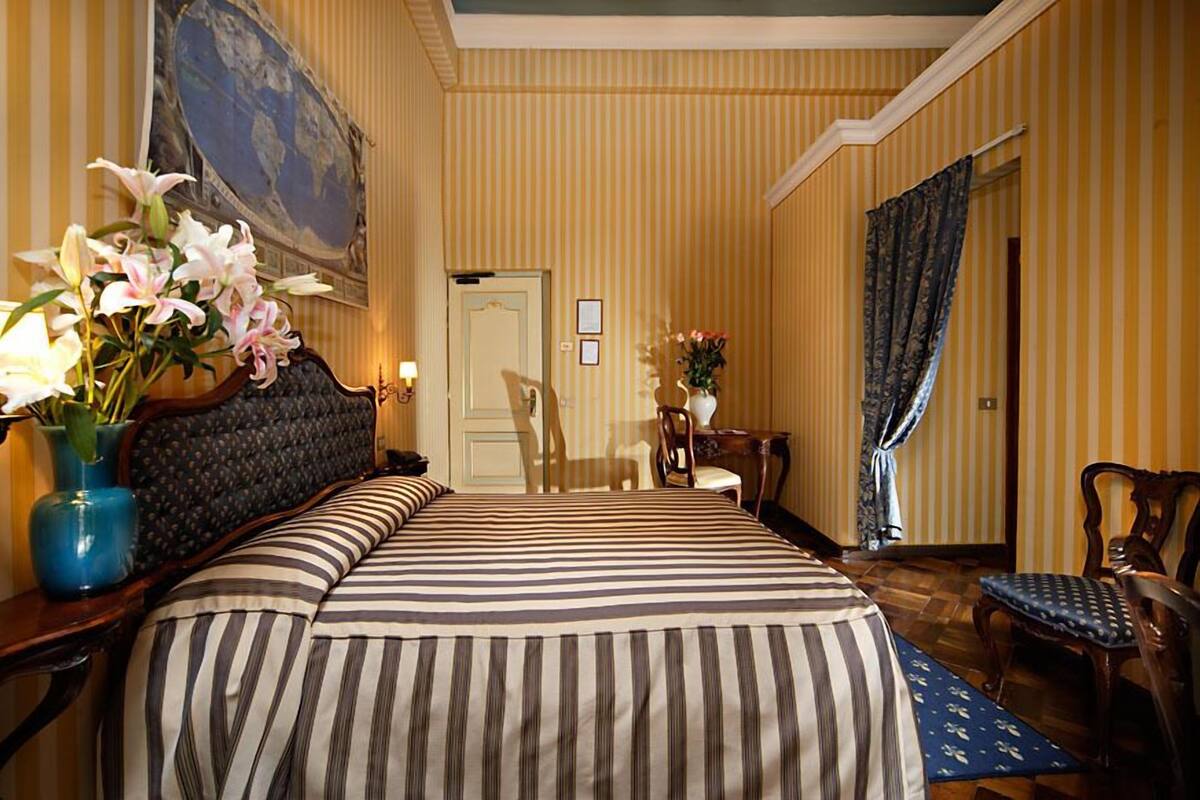 Quarto de hotel com uma cama grande, roupa de cama listrada, um vaso de flores e vista para o corredor.