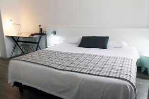 Uma cama bem arrumada com colcha xadrez, uma mesa de cabeceira com abajur e uma cadeira.