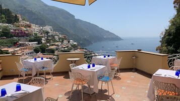 Terraço ao ar livre com mesas e cadeiras, com vista para uma cidade costeira e o mar.