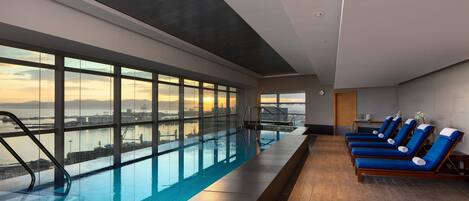 Piscina interna moderna com vista para a cidade, piso de madeira e espreguiçadeiras.