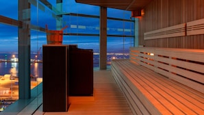 Quarto de hotel moderno com grandes janelas, um banco de madeira e vista para a cidade ao entardecer.